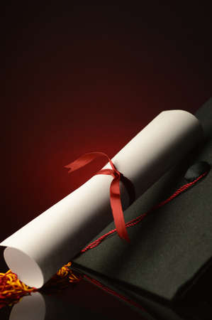 A closeup view of a diploma and mortarboard grad hat over a dark red ...