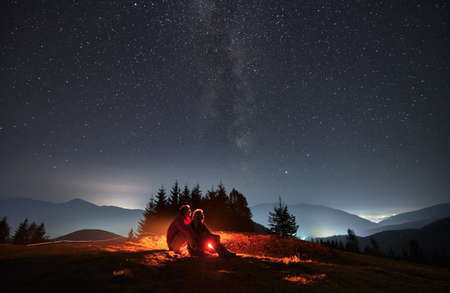Young man and woman travelers sitting on grassy hill under night sky ...