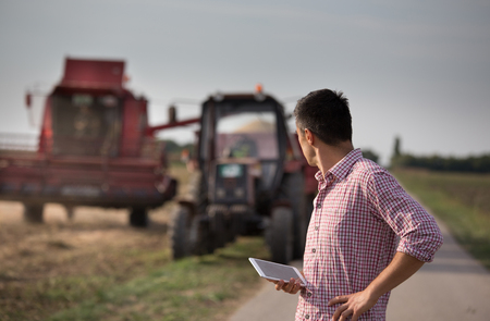 Handsome farmer with tablet standing in front of combine and tractor ...