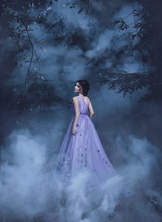 Beautiful young brunette in a luxurious purple long dress. Fairy Photo. Creative toning. Queen walking in the fog. FY31064597610