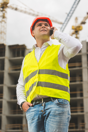 Young smiling engineer in hardhat and safety vest talking by phone on ...
