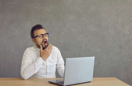 Sleep deprived young man in glasses sitting at office desk and yawning ...