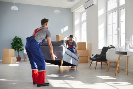 Happy young workers from the moving company lift up a heavy couch. Two ...