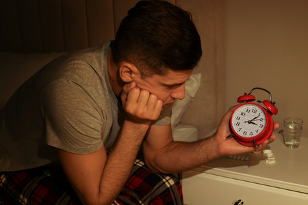 Man suffering from insomnia looking at time on alarm clock in bedroom ...