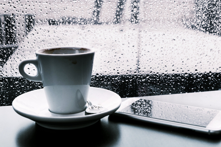 closeup of a cup of coffee and a smartphone on a table while is raining ...