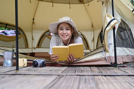 Cheerful young woman lying in her camping tent and reading a book ...