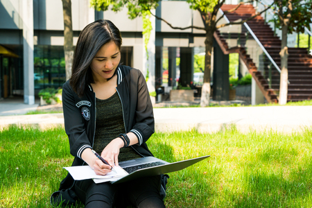 A young student woman is writing note while looking at laptop computer ...