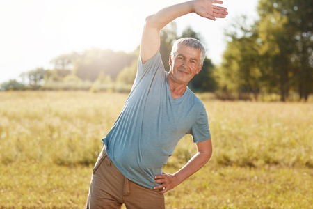 Outdoor shot of active senior male pensioner bends body, does physical ...