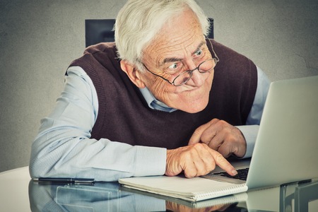 Elderly old man using computer sitting at table isolated on grey wall ...