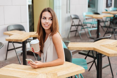 Smiling young woman using cell phone and drinking coffee in outdoor ...