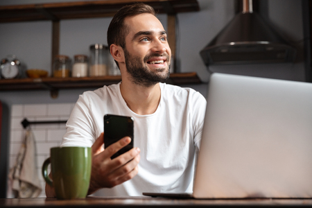 Happy young man using laptop computer while sitting at the kitchen ...