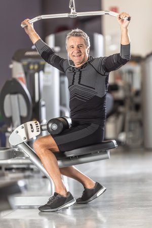Young at heart male pensioner pulls weights in a gym with headphones on ...