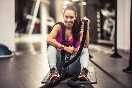 Woman holding battle ropes sits on a mat nside the gym, smiling at the ...